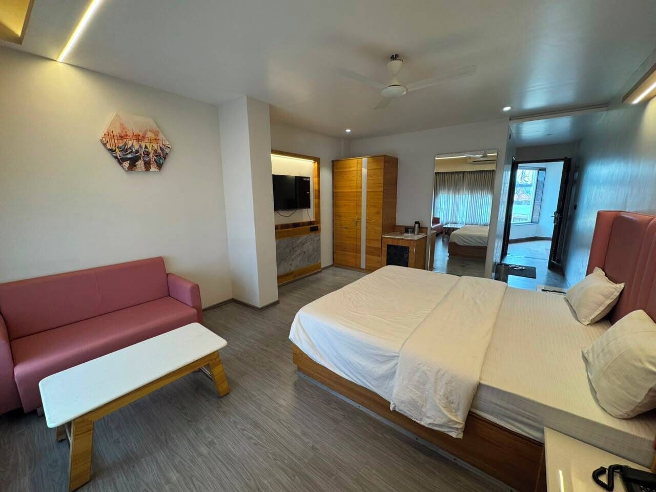 Quarto de hotel moderno com cama, sofá, uma mesinha, televisão e banheiro visível por uma porta aberta.