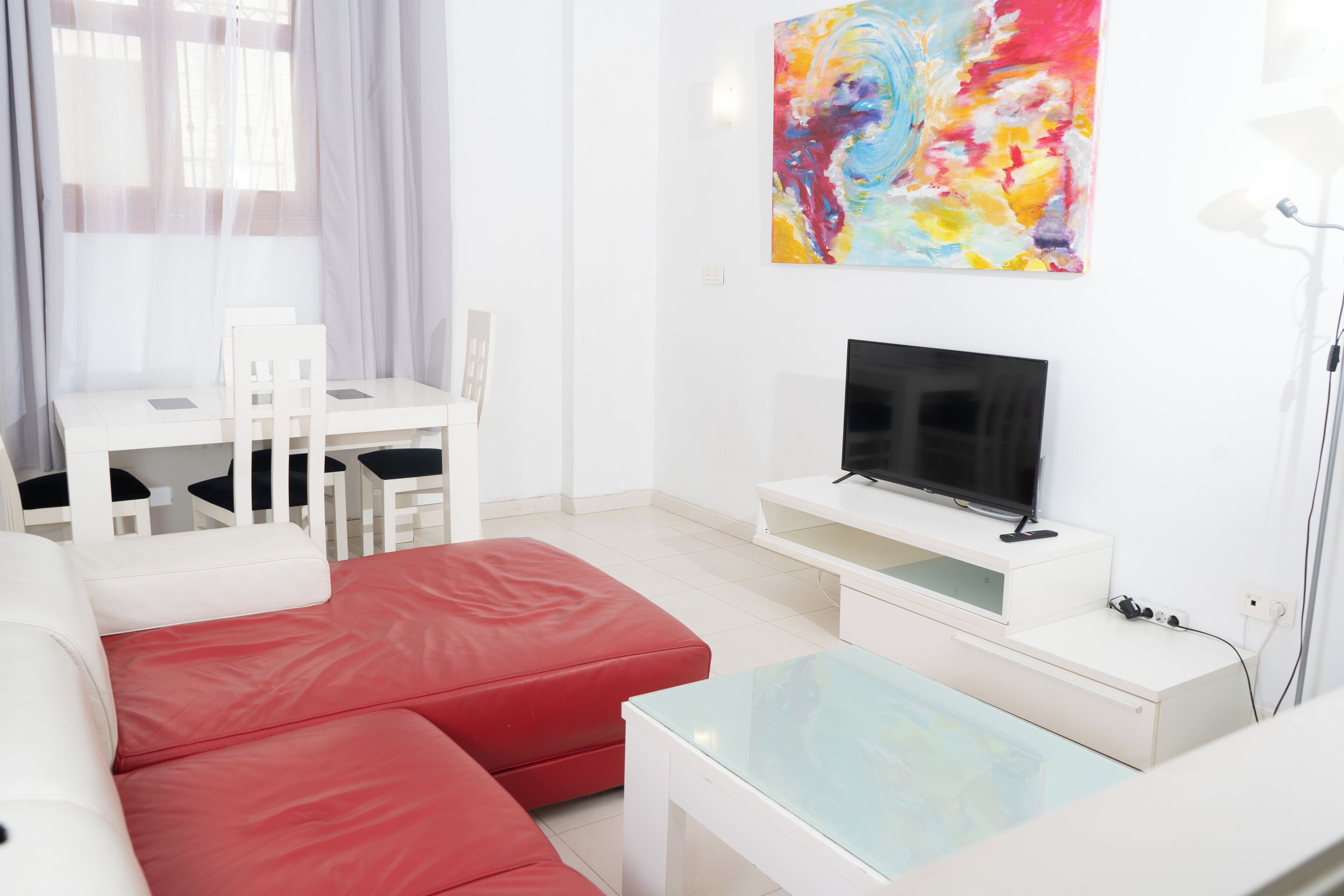 Uma sala de estar moderna com um sofá branco, um banquinho vermelho, uma mesa de centro de vidro, uma TV sobre um suporte branco e um quadro abstrato colorido na parede.
