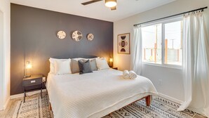 Um quarto com uma cama, mesa de cabeceira, ventilador de teto, obras de arte na parede e uma janela com cortinas.