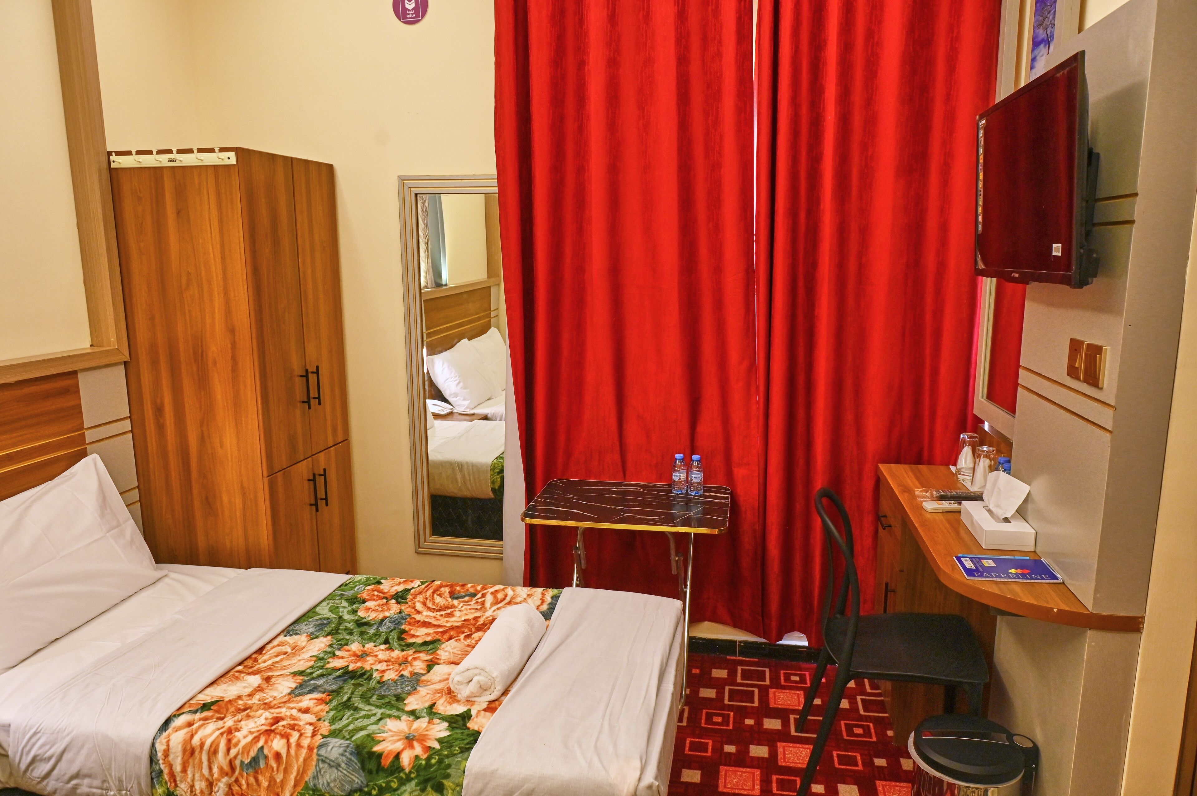 Quarto de hotel com uma cama, um guarda-roupa de madeira, uma mesinha com um espelho, uma escrivaninha com uma cadeira e uma cortina vermelha.
