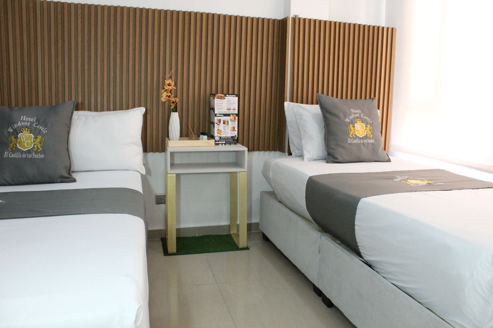 Quarto de hotel com duas camas, uma mesinha e cabeceira de madeira.