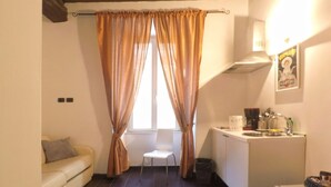 Um quarto pequeno e acolhedor com sofá, uma cadeira, cozinha compacta e janela com cortinas.