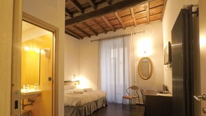 Quarto de hotel com cama, escrivaninha, cadeira, espelho e banheiro com pia e vaso sanitário.