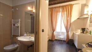 Casa de banho com lavatório, bidé e espelho. Tem uma janela com cortinas, uma cadeira e uma pequena mesa com uma chaleira e chávenas.