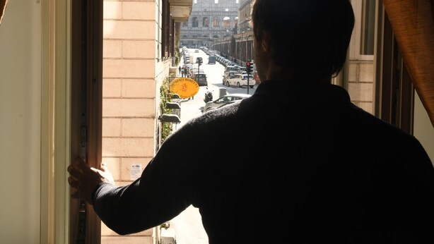 Uma pessoa olhando pela janela para uma rua com edifícios e carros estacionados.