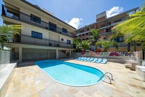 Piscina com espreguiçadeiras, área coberta e um edifício de vários andares com varandas.