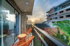 Uma varanda com cadeiras de vime, uma porta de vidro e vista para o pôr do sol sobre edifícios.