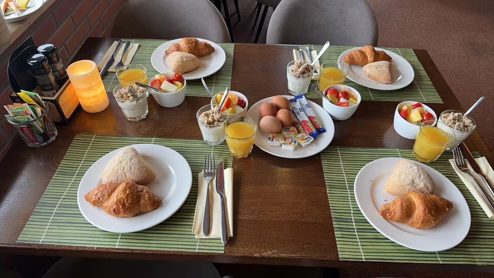 Mesa de café da manhã com pratos de croissants, ovos e frutas, acompanhada de suco e cereal.