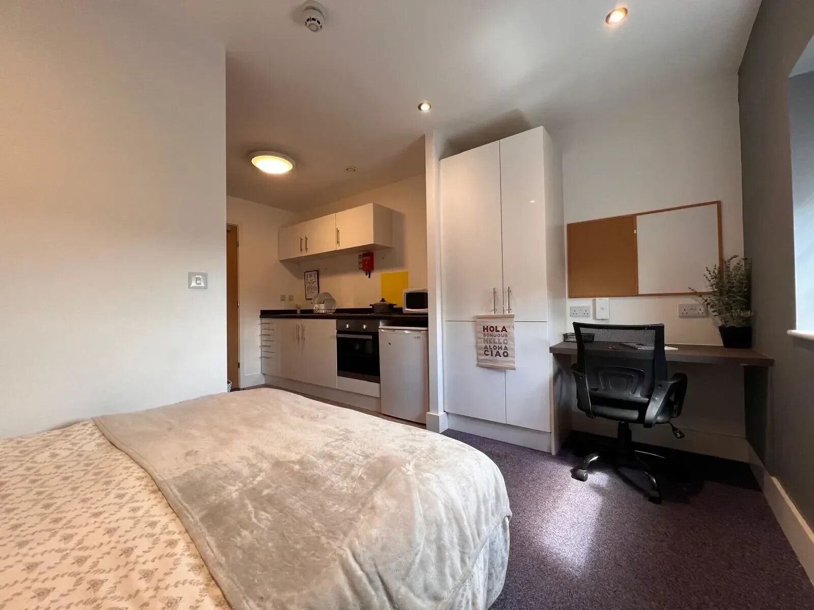 Quarto compacto com uma cama, uma mesa com cadeira, uma cozinha compacta e um aviso com saudações em vários idiomas.