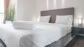 Uma cama bem arrumada com lençóis e almofadas brancas, uma mesa de cabeceira com abajur e um vaso de planta perto da janela.