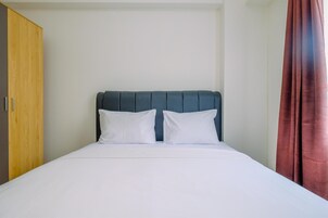 Uma cama com cabeceira escura, roupa de cama branca e um guarda-roupa de madeira à esquerda.