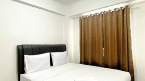 Cama bem arrumada com roupa de cama branca e toalha branca dobrada, cabeceira preta e cortina marrom.