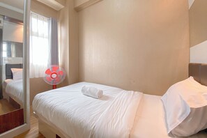 Quarto de hotel com uma cama de solteiro, um ventilador portátil e uma janela com cortinas.