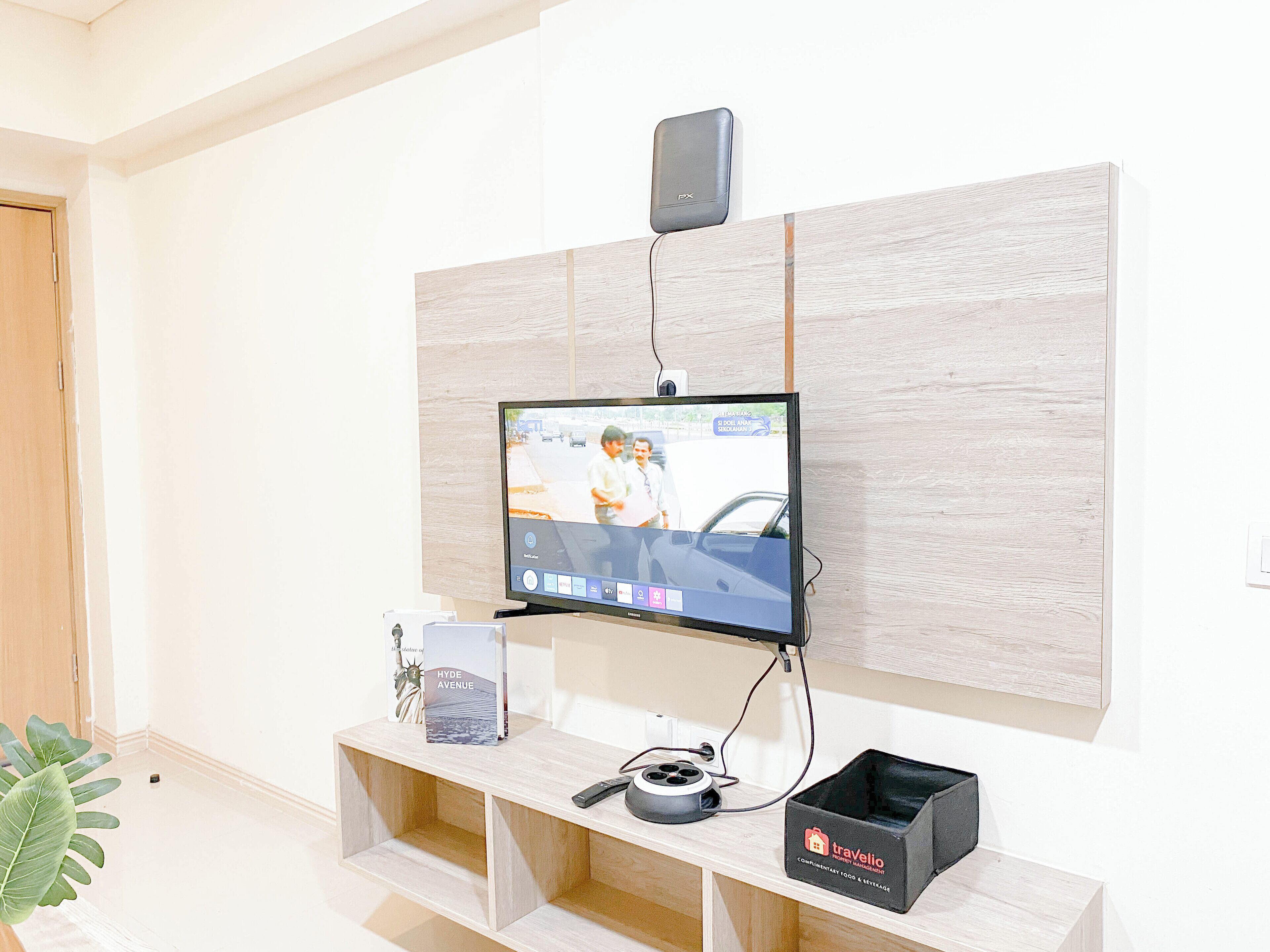 Uma TV de tela plana fixada na parede, com um painel de madeira ao fundo, uma pequena prateleira abaixo com um controle remoto, um pequeno alto-falante e uma caixa rotulada 'Travelio'.