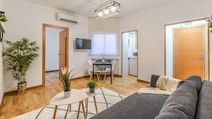 Uma sala de estar moderna com um sofá cinzento, uma pequena mesa de jantar com cadeiras, uma televisão na parede e uma porta de madeira.