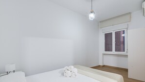 Quarto minimalista com uma cama de solteiro, mesa de cabeceira com abajur, janela com persianas e luminária pendente.