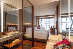 Quarto de hotel moderno com uma cama grande, banheiro separado com pia e espelho, e vista para a cidade através de amplas janelas.