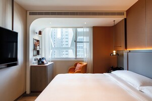 Quarto de hotel com uma cama grande, televisão, escrivaninha com cadeira, janela com vista para edifícios e abajur.