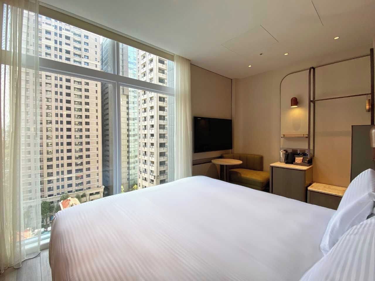 Quarto de hotel com uma cama grande, televisão, mesa pequena, cadeira e vista para edifícios altos.