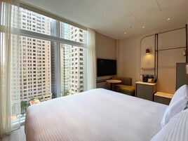 Quarto de hotel com uma cama grande, televisão, mesa pequena, cadeira e vista para edifícios altos.