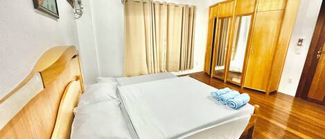 Quarto de hotel com cama, cabeceira de madeira, cortinas bege, ventoinha no teto e guarda-roupa.