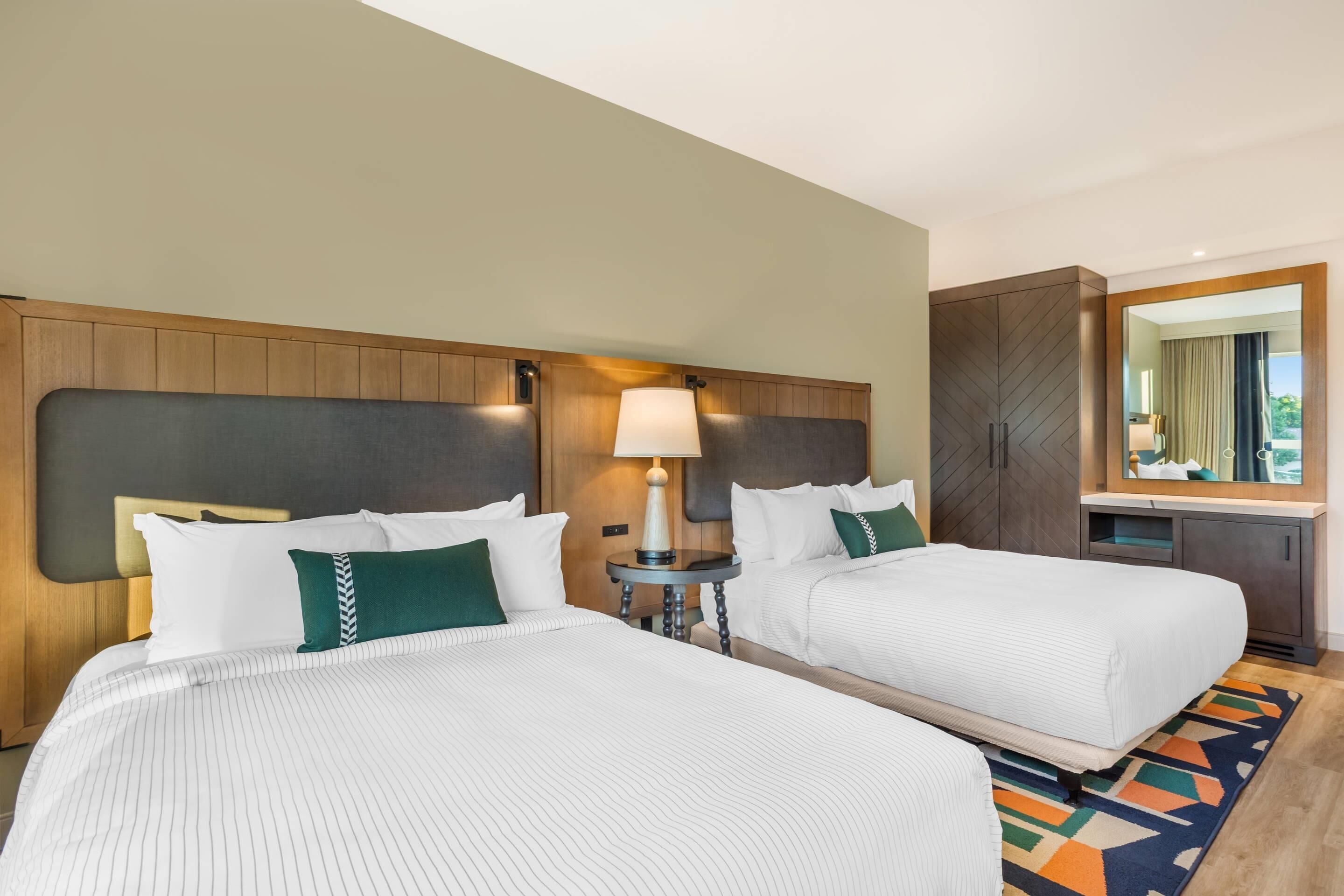 Quarto de hotel com duas camas, mesa de cabeceira com abajur, espelho e cabeceira de madeira.