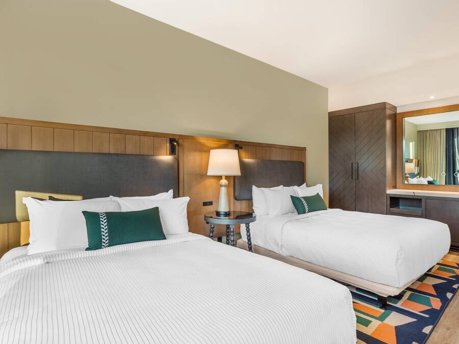 Quarto de hotel com duas camas, mesa de cabeceira com abajur, espelho e cabeceira de madeira.