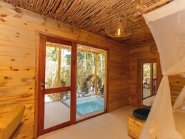 Uma cabana de madeira com porta de vidro que dá para uma área de piscina, uma cama com dossel e um lustre pendurado.