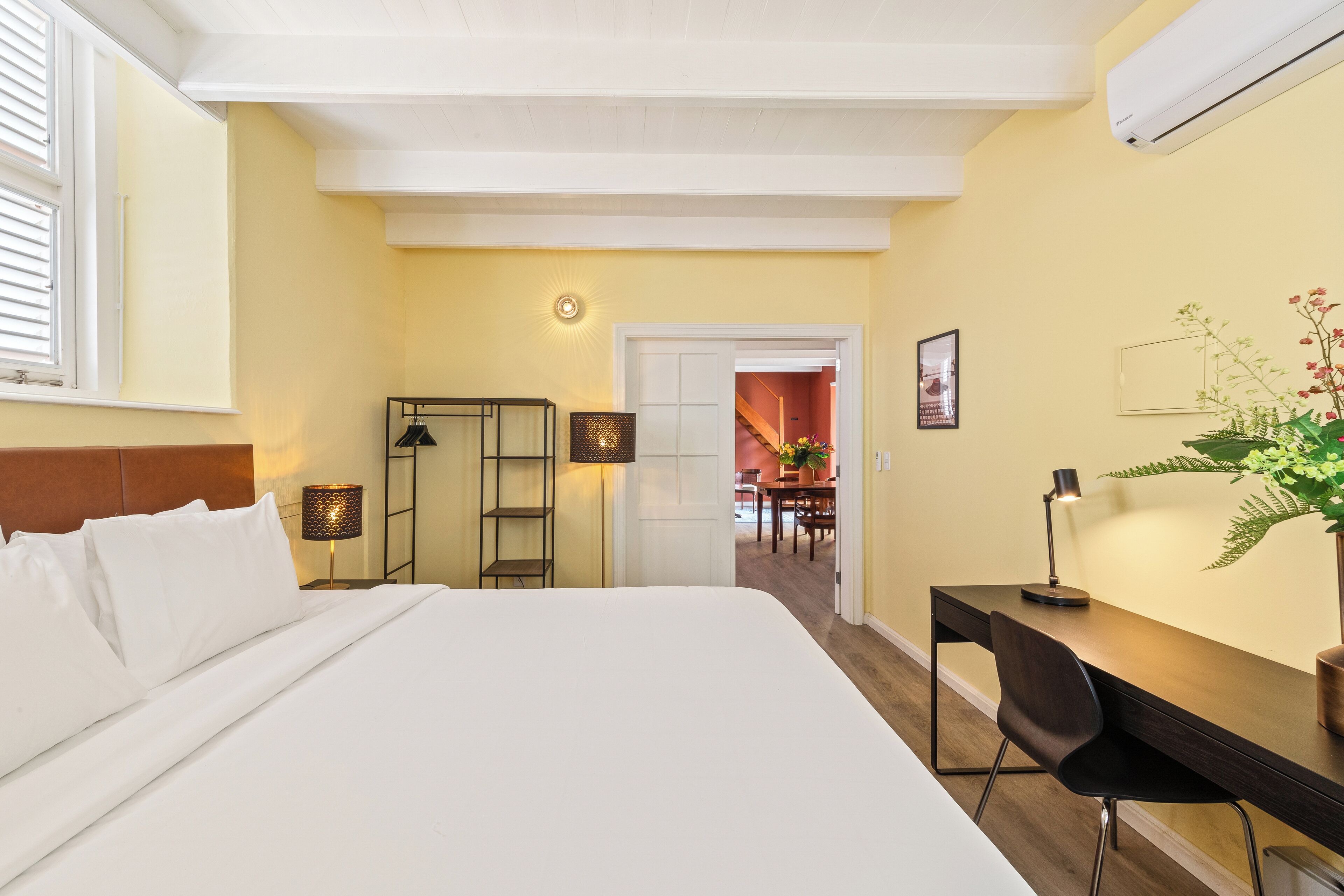 Quarto de hotel com cama, escrivaninha, cadeira, luminária e planta em vaso.