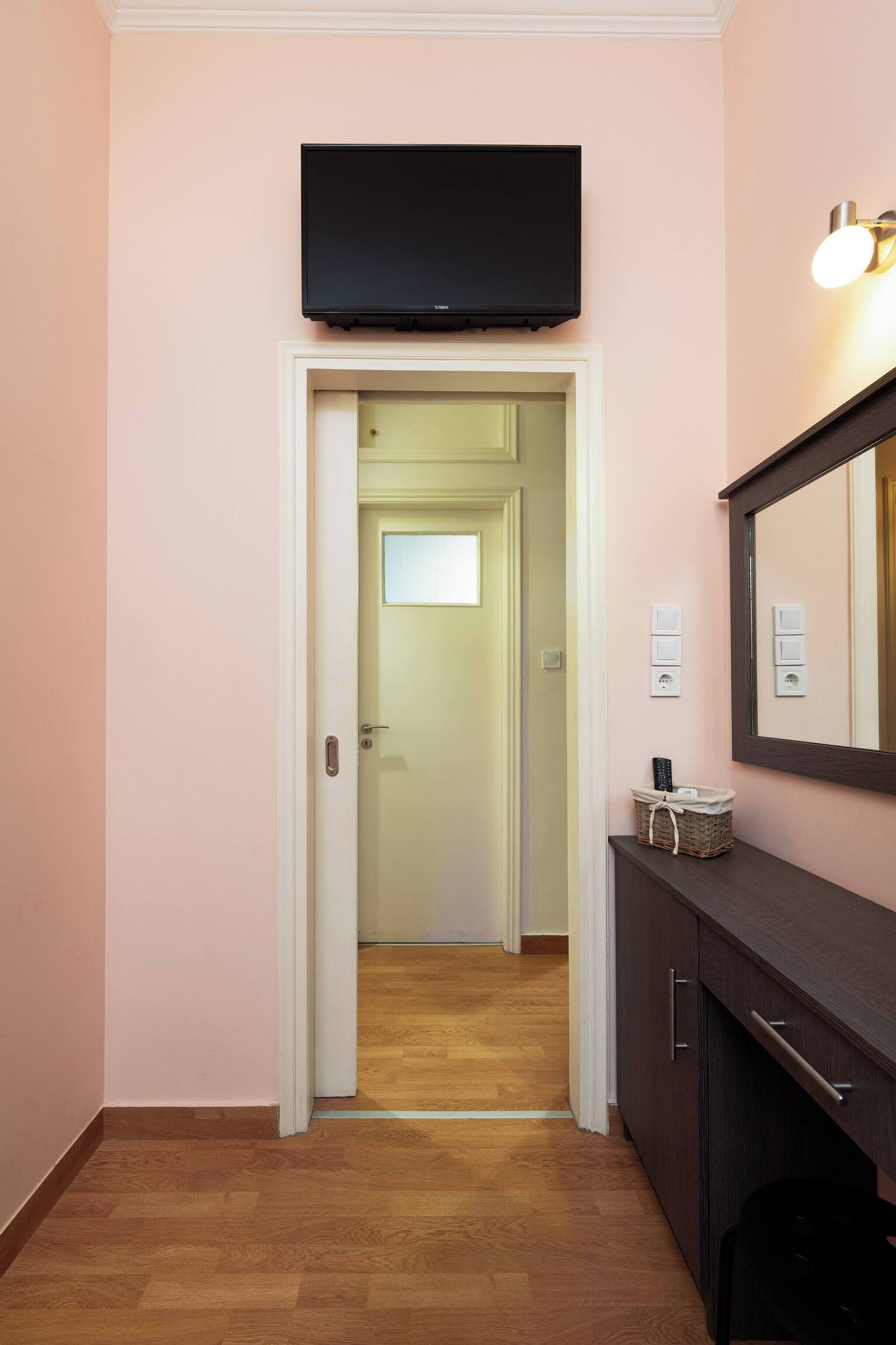 Um corredor com piso de madeira, uma televisão montada acima de uma porta, um espelho na parede e uma cesta sobre um balcão.