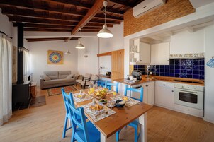 Uma sala de jantar com mesa de madeira posta para o café da manhã, cadeiras azuis e uma cozinha com azulejos azuis e armários brancos.