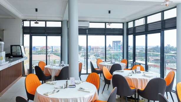 Restaurante com mesas redondas e cadeiras laranja, janelas amplas com vista para a cidade e um balcão com máquina de café.
