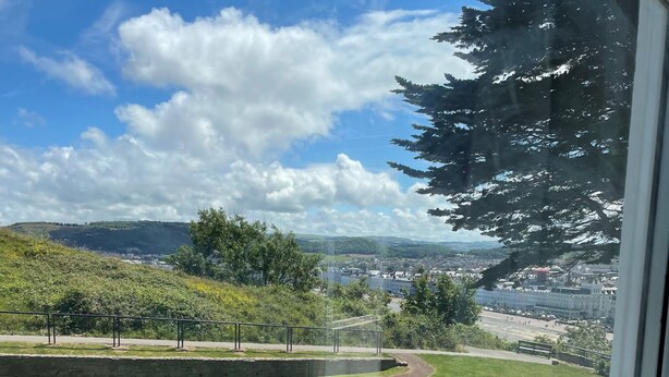 Vista de uma área gramada com um caminho, árvores e uma cidade ao longe.