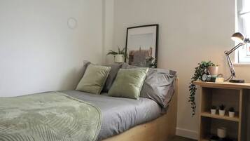 Quarto minimalista com cama, abajur, prateleira com plantas e quadro pendurado na parede.