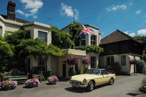 Um carro amarelo clássico estacionado em frente a um edifício com a bandeira britânica, rodeado por vegetação exuberante e flores.