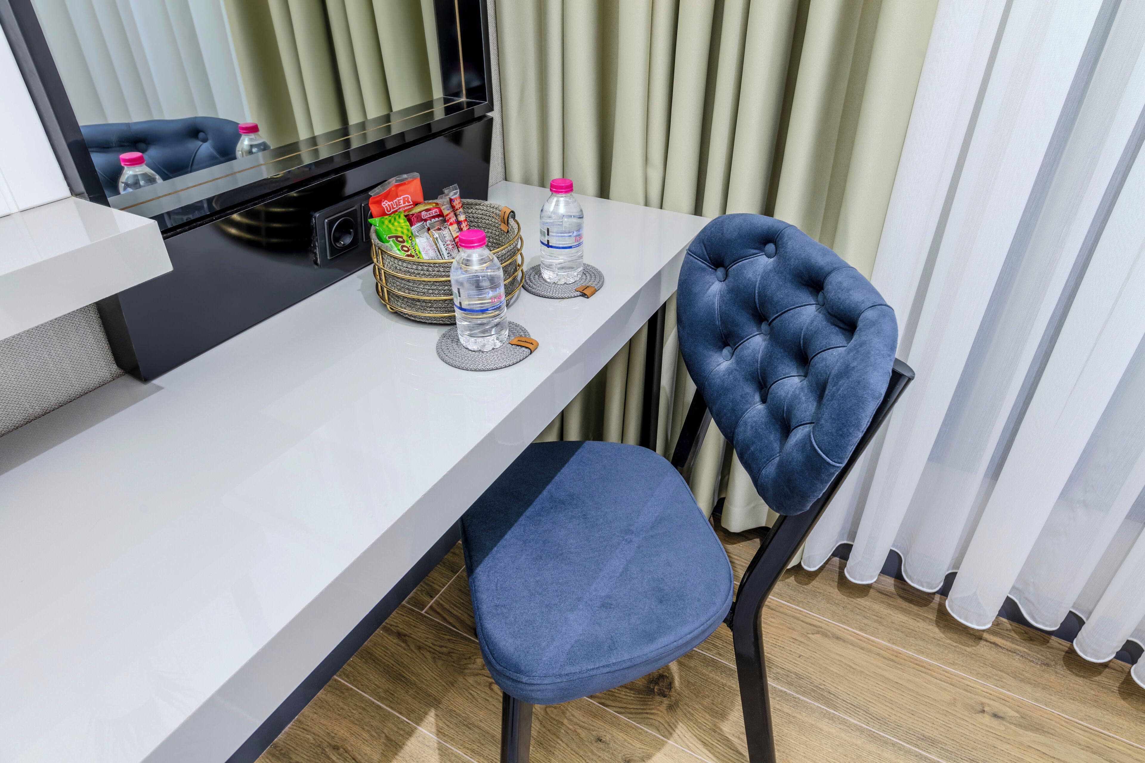 Quarto de hotel moderno com escrivaninha branca, poltrona azul estofada, cesta de vime com lanches e duas garrafas de água.
