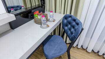 Quarto de hotel moderno com escrivaninha branca, poltrona azul estofada, cesta de vime com lanches e duas garrafas de água.
