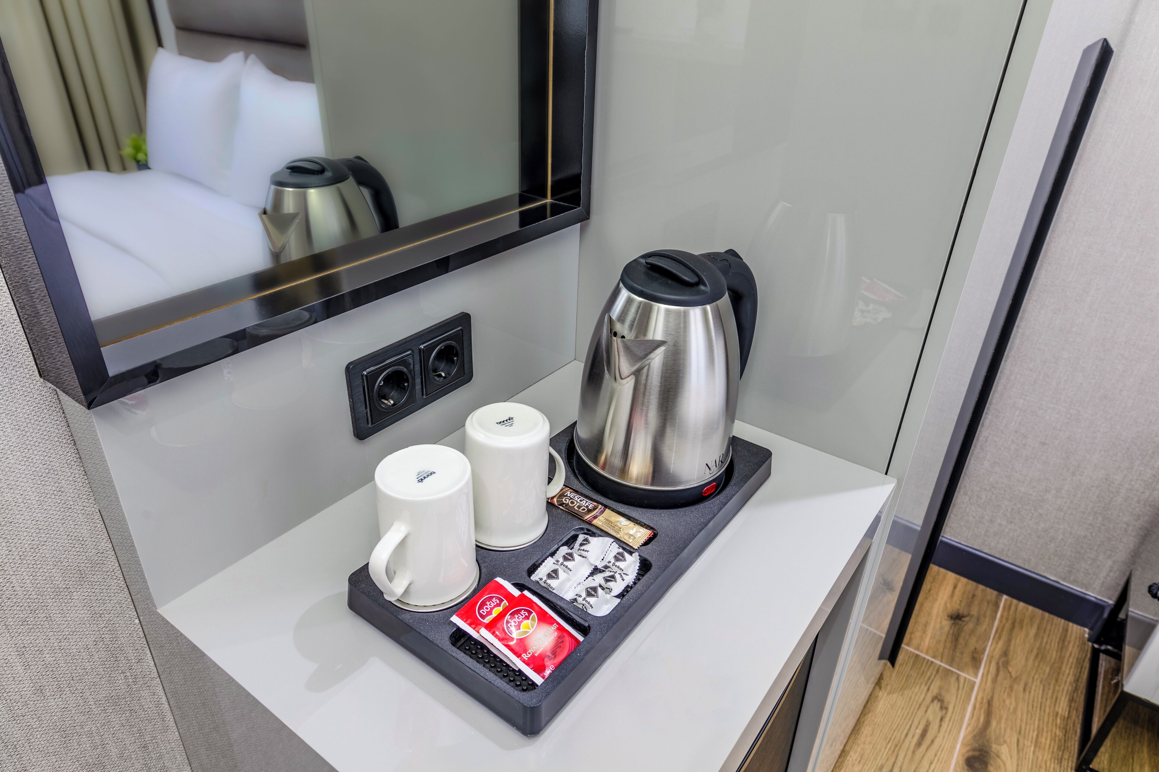 Quarto de hotel com minibar contendo uma chaleira, duas canecas brancas, uma caixa de saquinhos de chá e um pacote de café.