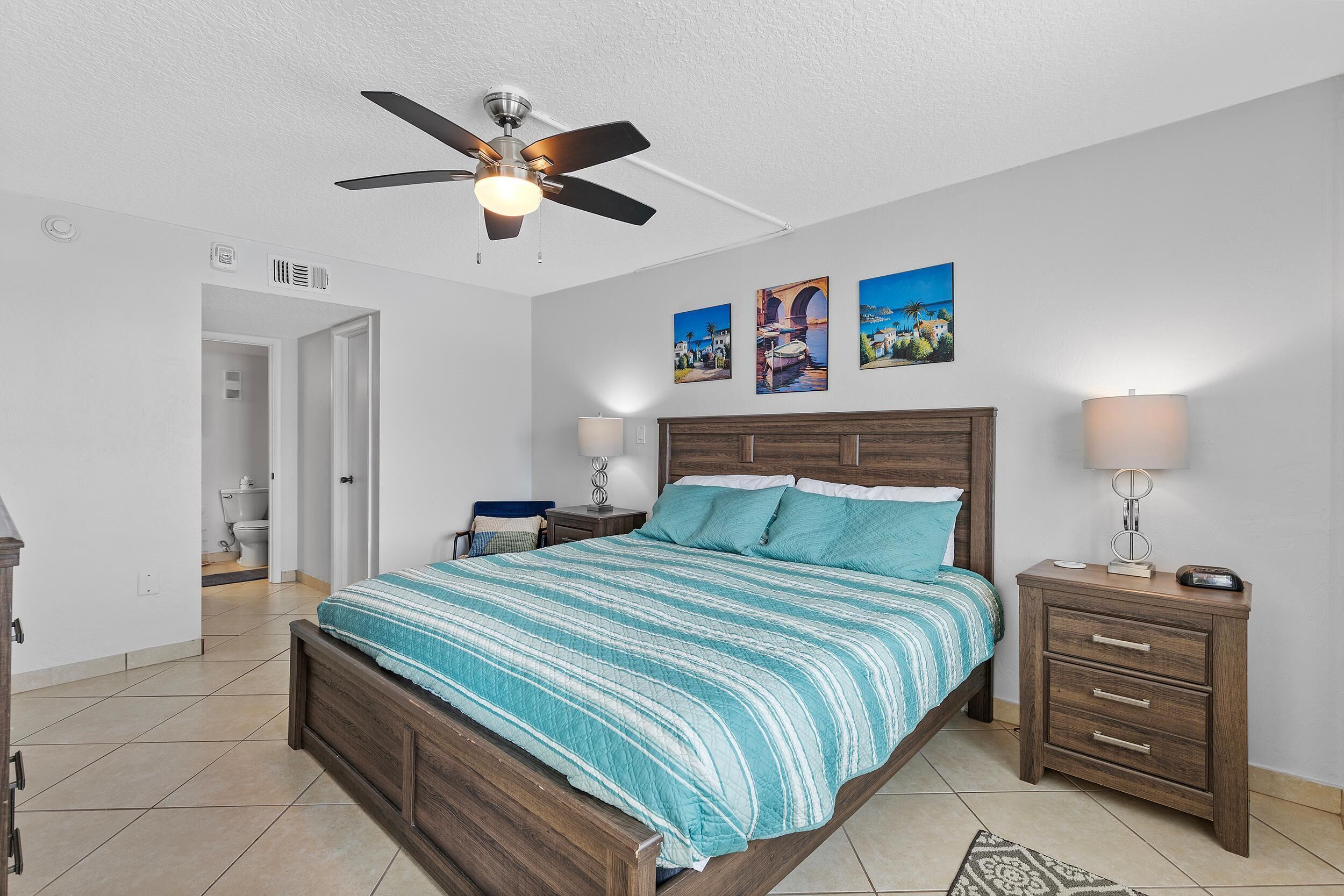 Quarto com cama, mesa de cabeceira, abajur, ventilador de teto e porta que dá para outro cômodo.