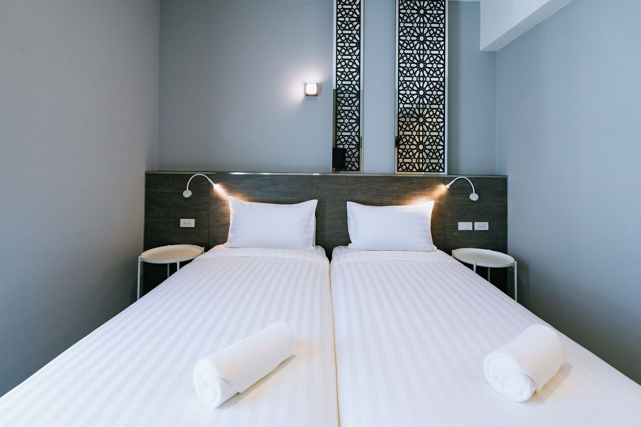 Cama de casal com lençóis brancos e duas almofadas, cabeceira com iluminação embutida, painel decorativo na parede e duas mesas de cabeceira.