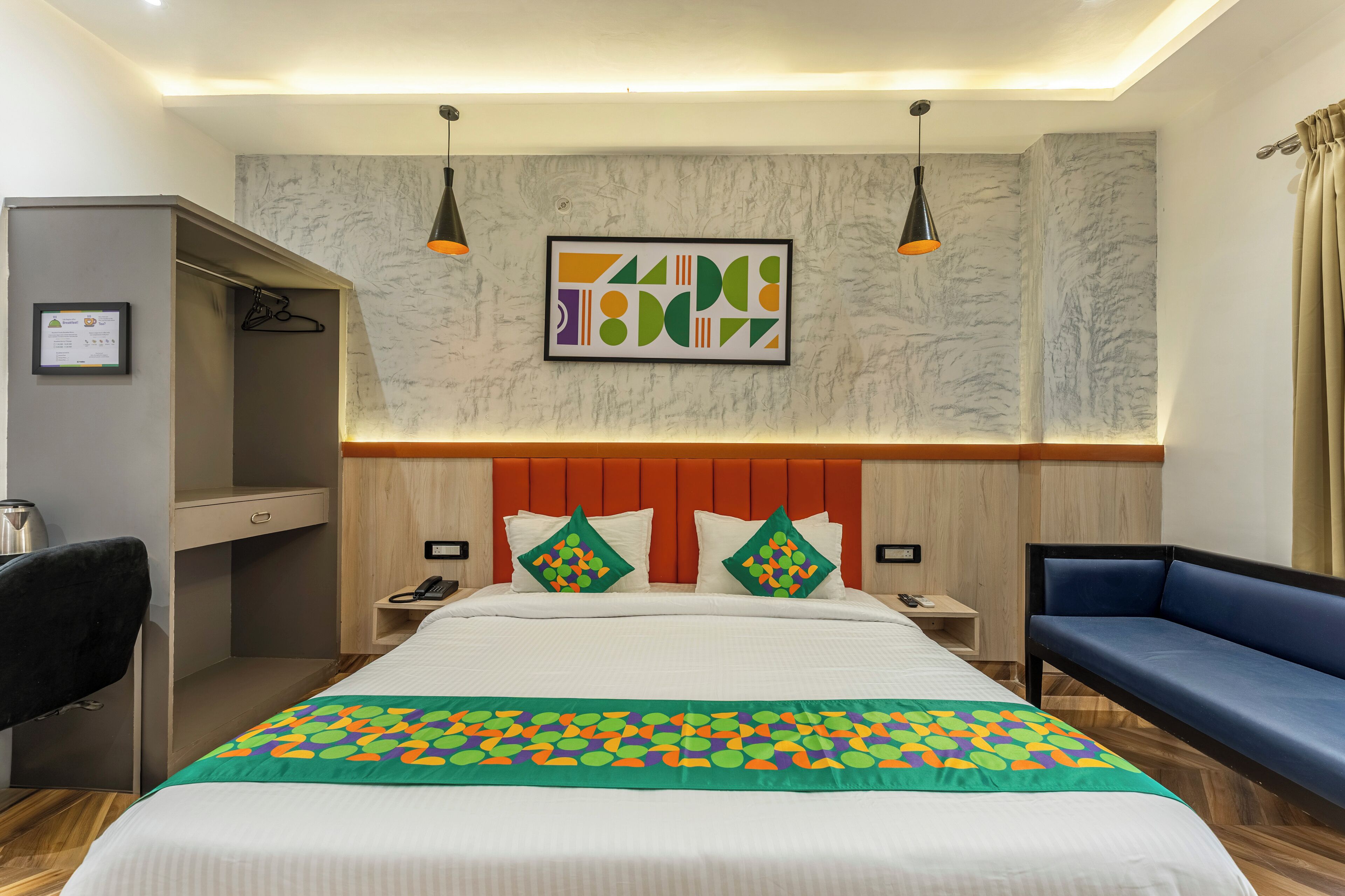 Quarto de hotel com cama, escrivaninha, cadeira, sofá azul, obra de arte na parede e mesa de cabeceira com telefone.