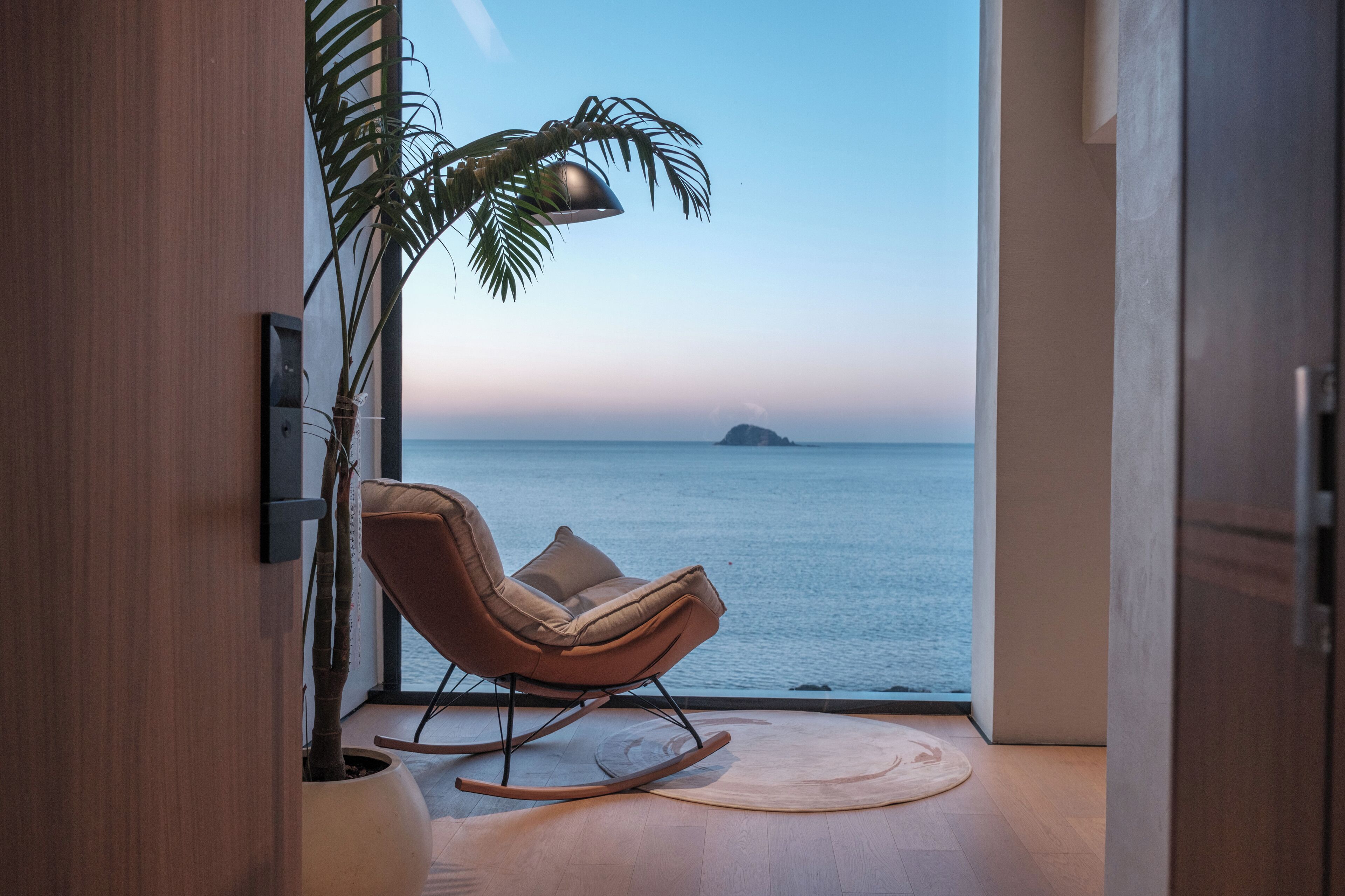Quarto com poltrona de balanço voltada para uma grande janela com vista para o mar.