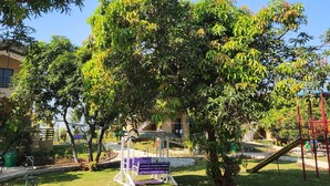 Uma área de parque com um parquinho, incluindo baloiços e um banco, rodeada por árvores e um caminho pavimentado.