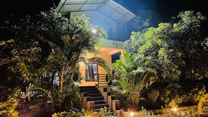 Uma casa moderna com teto de vidro, rodeada por vegetação exuberante e um jardim bem iluminado à noite.