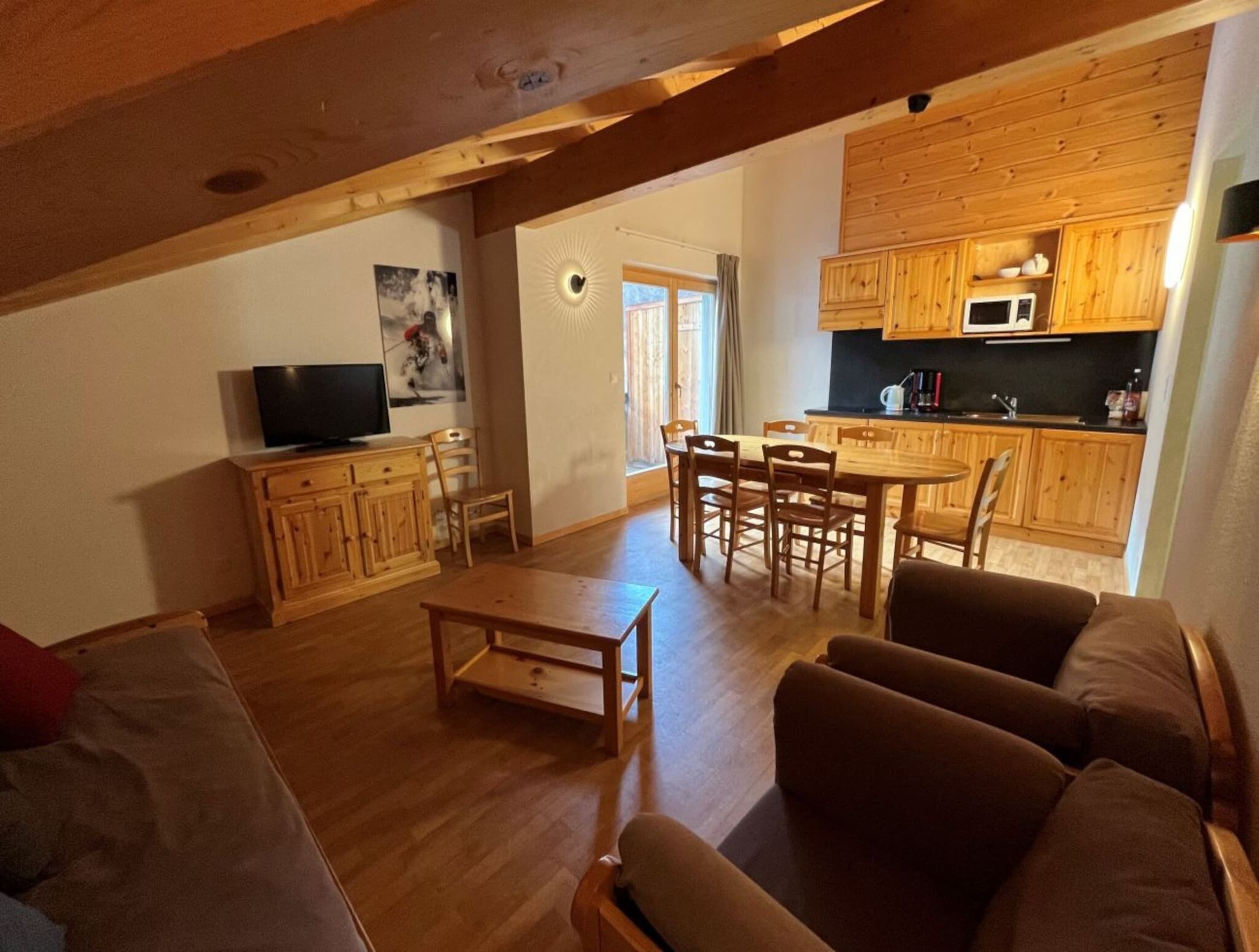 Uma sala de estar acolhedora, com piso de madeira, um sofá, uma mesa de centro e uma televisão. Há uma cozinha com armários de madeira e um micro-ondas.