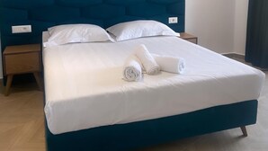 Uma cama bem arrumada com lençóis brancos e duas toalhas enroladas, um painel de cabeceira estofado em azul e uma mesa de cabeceira de madeira com tomada.