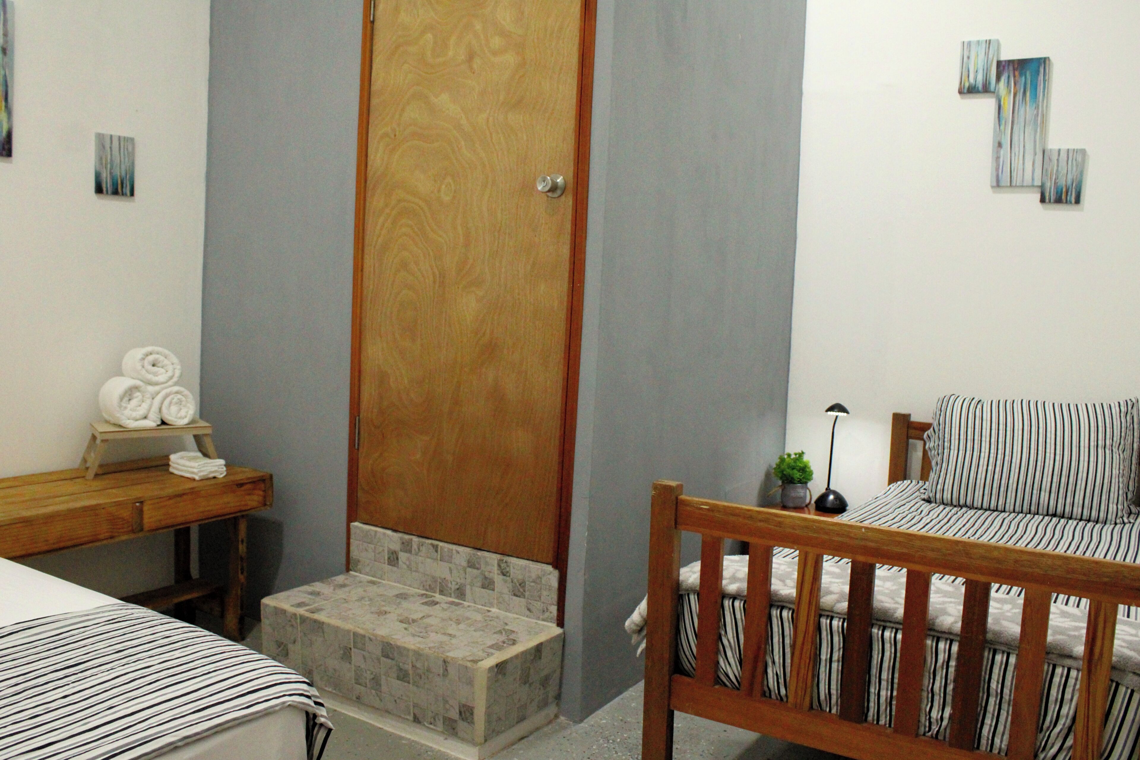 Quarto de hotel com porta de madeira, cama com roupa de cama listrada, mesa de cabeceira com toalhas e uma pequena planta.