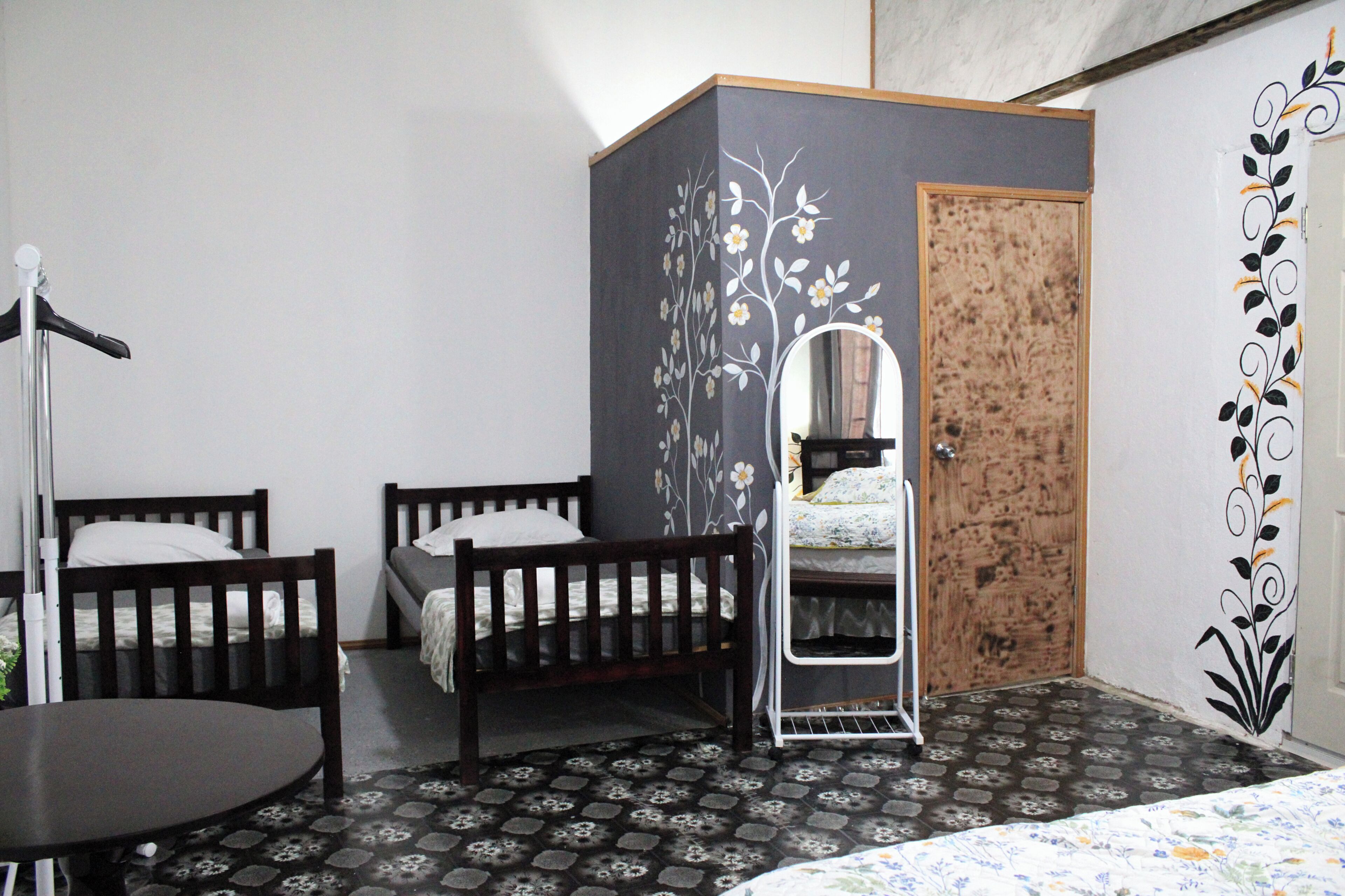 Quarto com duas camas de madeira, uma mesinha, um espelho e uma porta com papel de parede floral.
