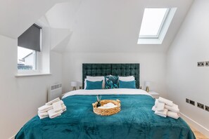 Quarto moderno com cama grande, roupa de cama azul-turquesa, cabeceira, claraboia e uma pequena mesa de cabeceira com luminária.