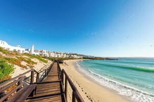 Um calçadão de madeira que leva a uma praia, com edifícios brancos ao fundo.